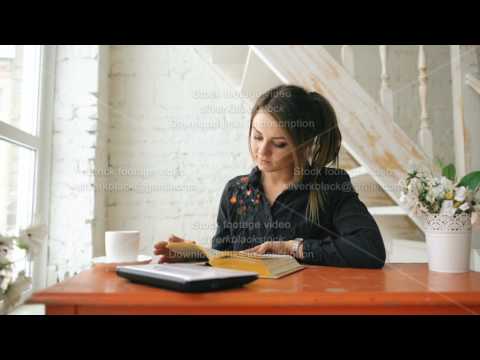 Young beautiful woman student sitting with laptop and reading book at cafe indoors