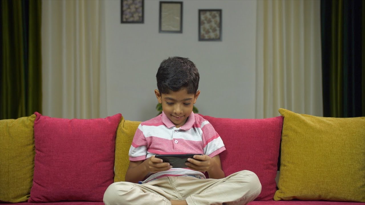Cute Indian kid playing mobile games while sitting on a couch ...