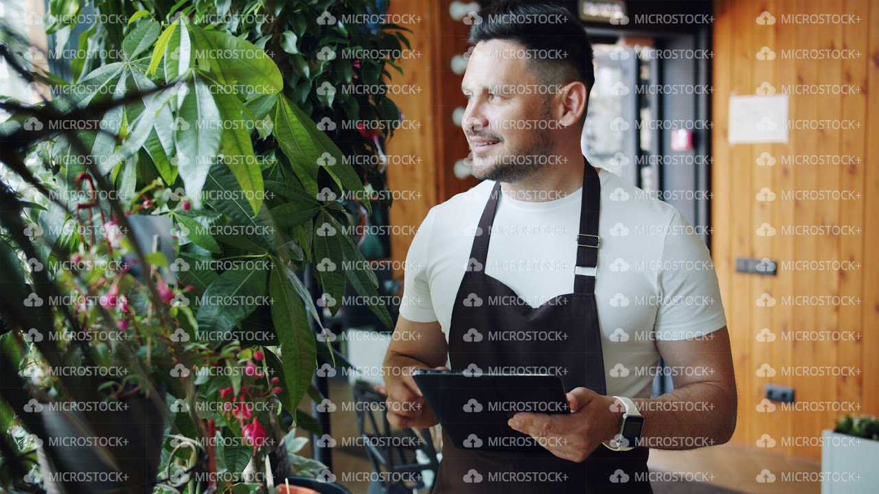 Flower shop manager touching plants smiling and using tablet in store ...