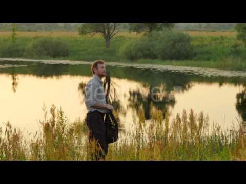 Man at Forest Lake Takes Off His Backpack Green Grass Field Smooth Water in a Pond Evening Dusk