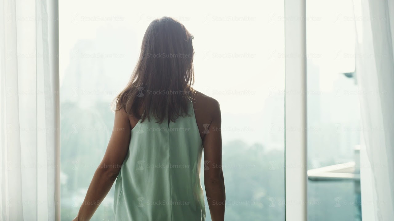 Young brunette woman opening curtains in morning. Girl looking through ...