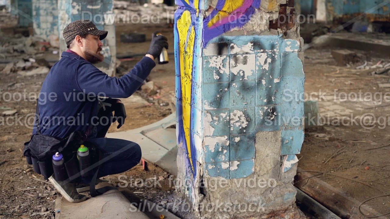 Modern graffiti painter is squatting near column in old abandoned ...