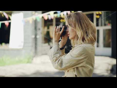 Cheerful young girl is adjusting camera then taking photos of beautiful city standing outdoors in