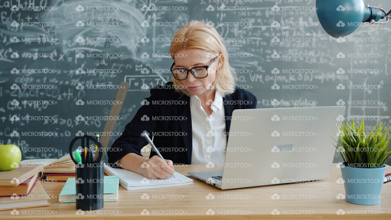 University professor using laptop then taking notes working at desk in ...